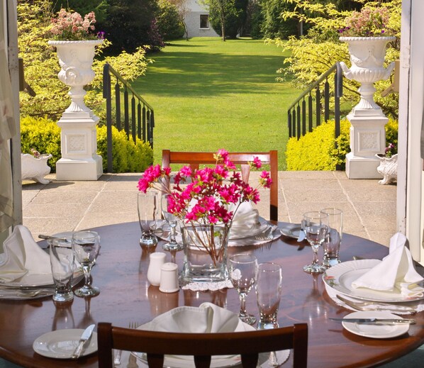 Mesa de jantar posta para seis pessoas, com flores cor-de-rosa, copos e talheres, com vista para um gramado e jardim bem cuidados.