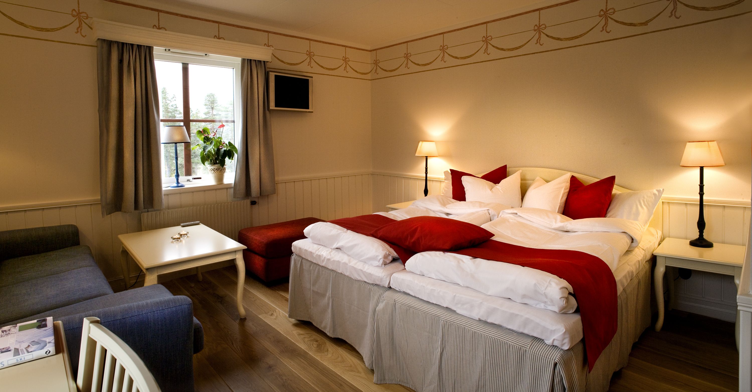 Quarto de hotel com cama, duas mesas de cabeceira com luminárias, uma mesinha, uma cadeira, janela com cortinas e uma televisão na parede.