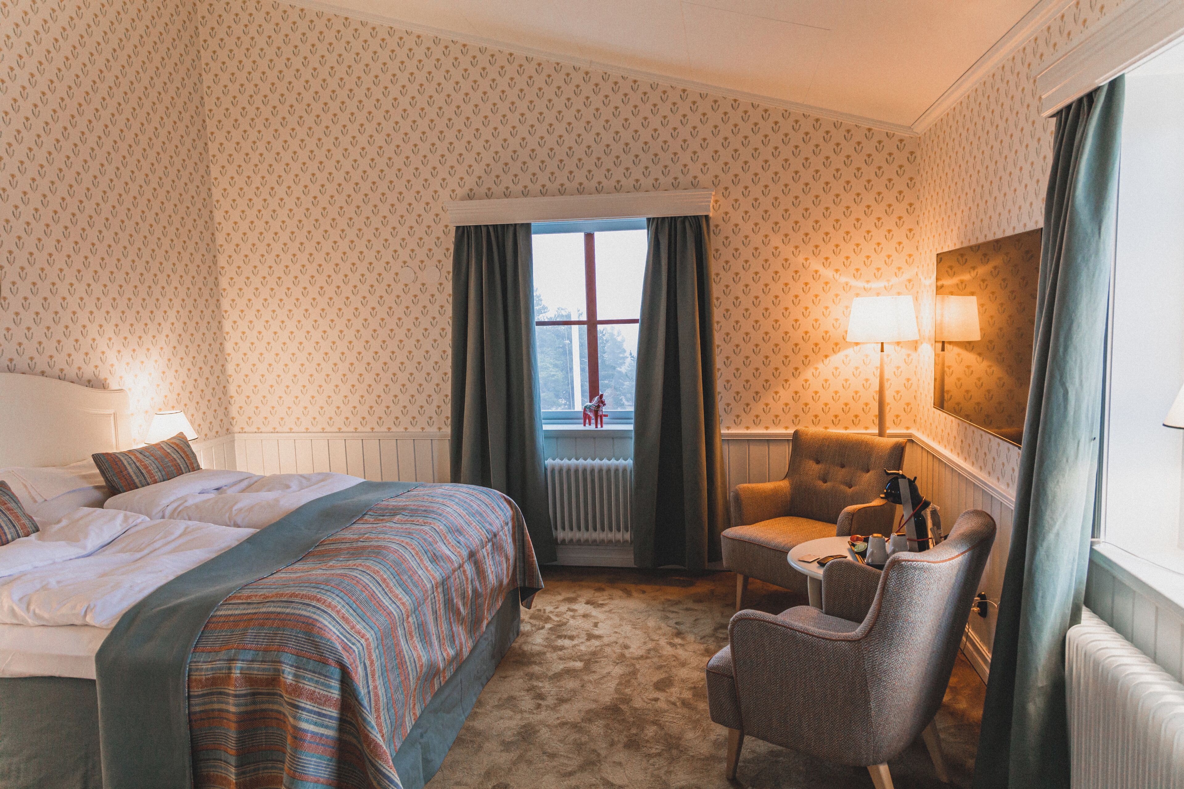 Quarto de hotel com uma cama, uma cadeira, uma mesinha, uma luminária e uma janela com cortinas.