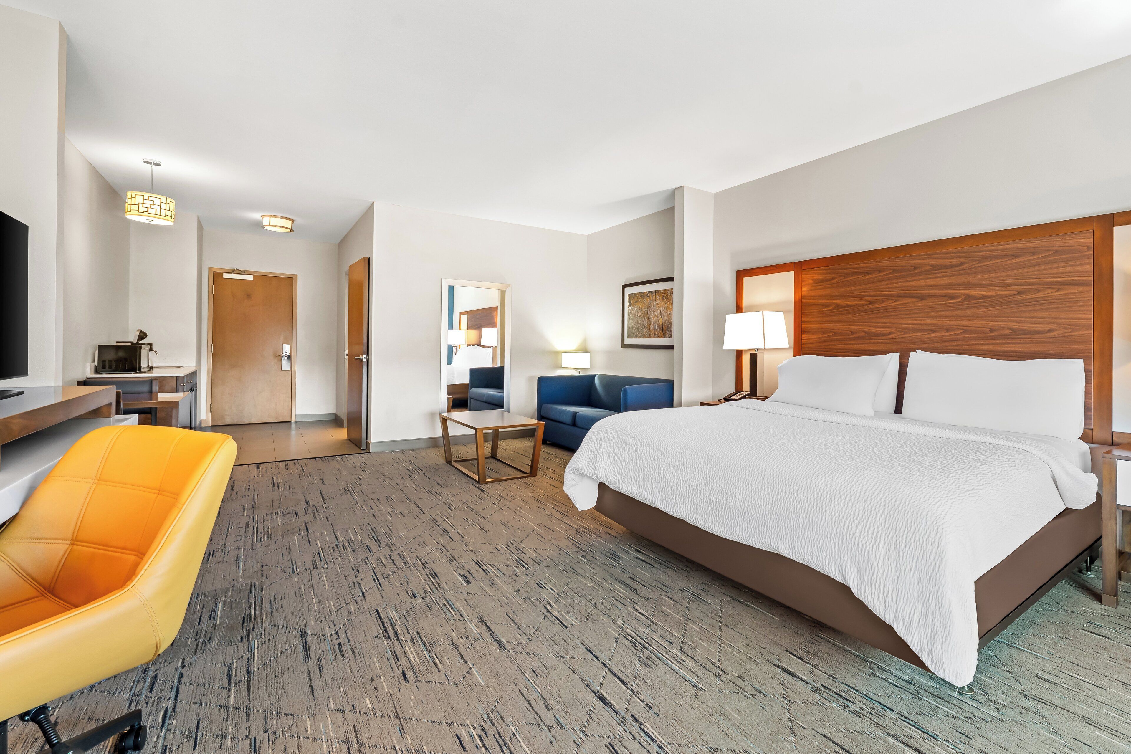 Quarto de hotel com uma cama grande, uma escrivaninha com cadeira, um sofá, uma televisão e uma porta que dá para outro quarto.
