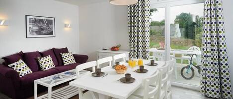 Uma sala de estar moderna com um sofá roxo, uma mesa de jantar branca e uma grande janela com cortinas.