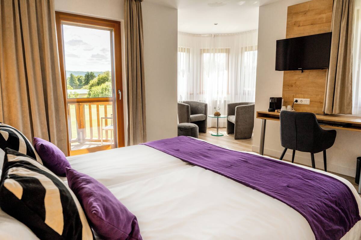Quarto de hotel com uma cama grande, uma escrivaninha com cadeira, televisão e varanda com vista.