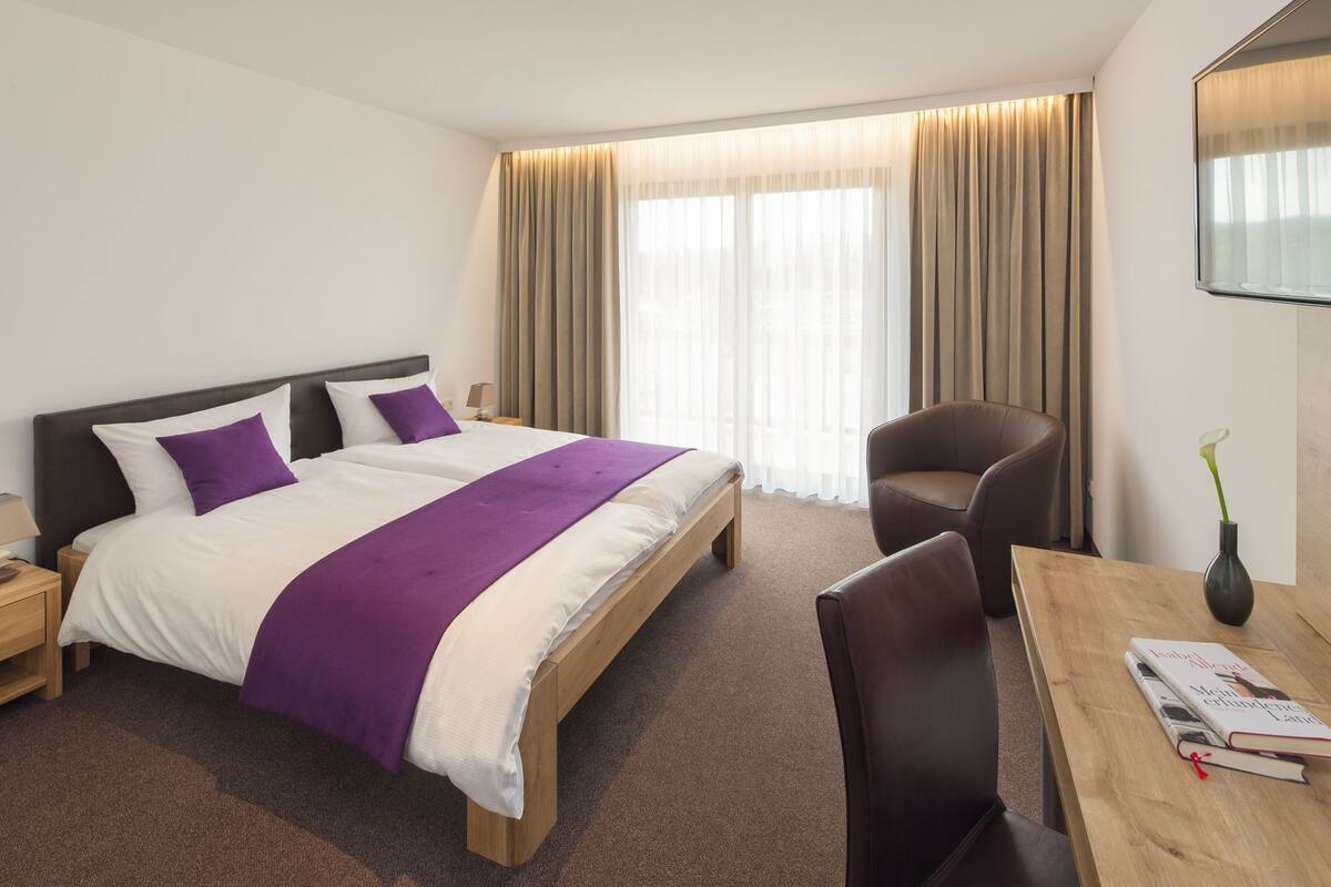 Quarto de hotel com cama, mesa de cabeceira de madeira, escrivaninha com cadeira, um vaso com planta e janela com cortinas.