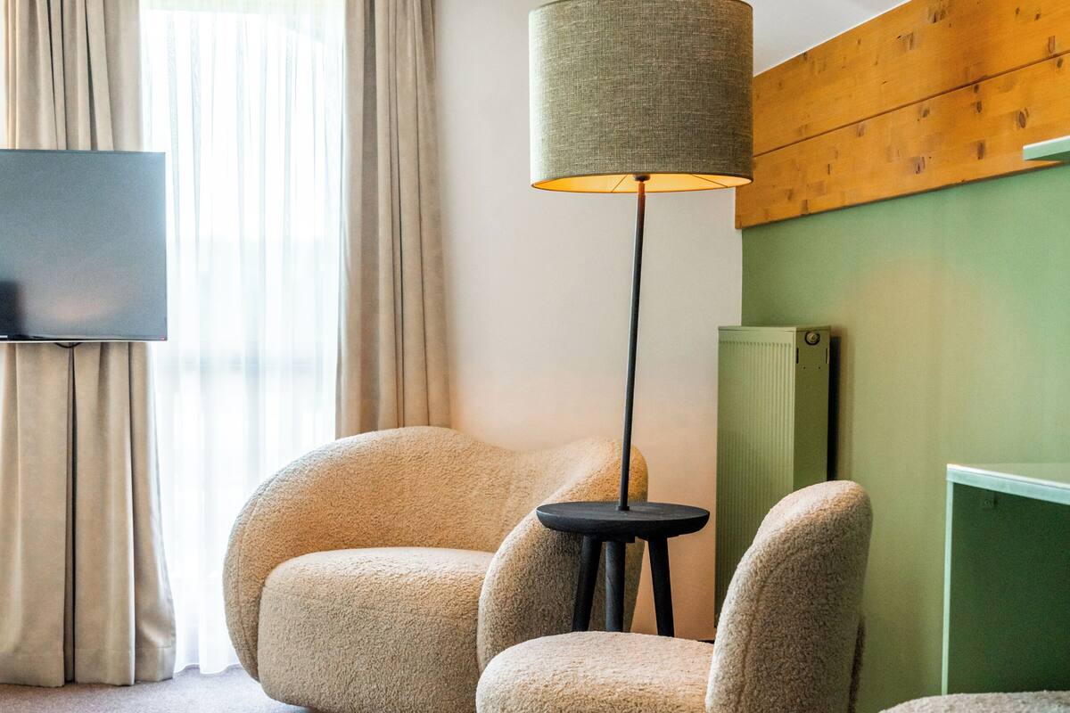 Quarto de hotel com duas poltronas bege, um abajur, uma televisão e um radiador.