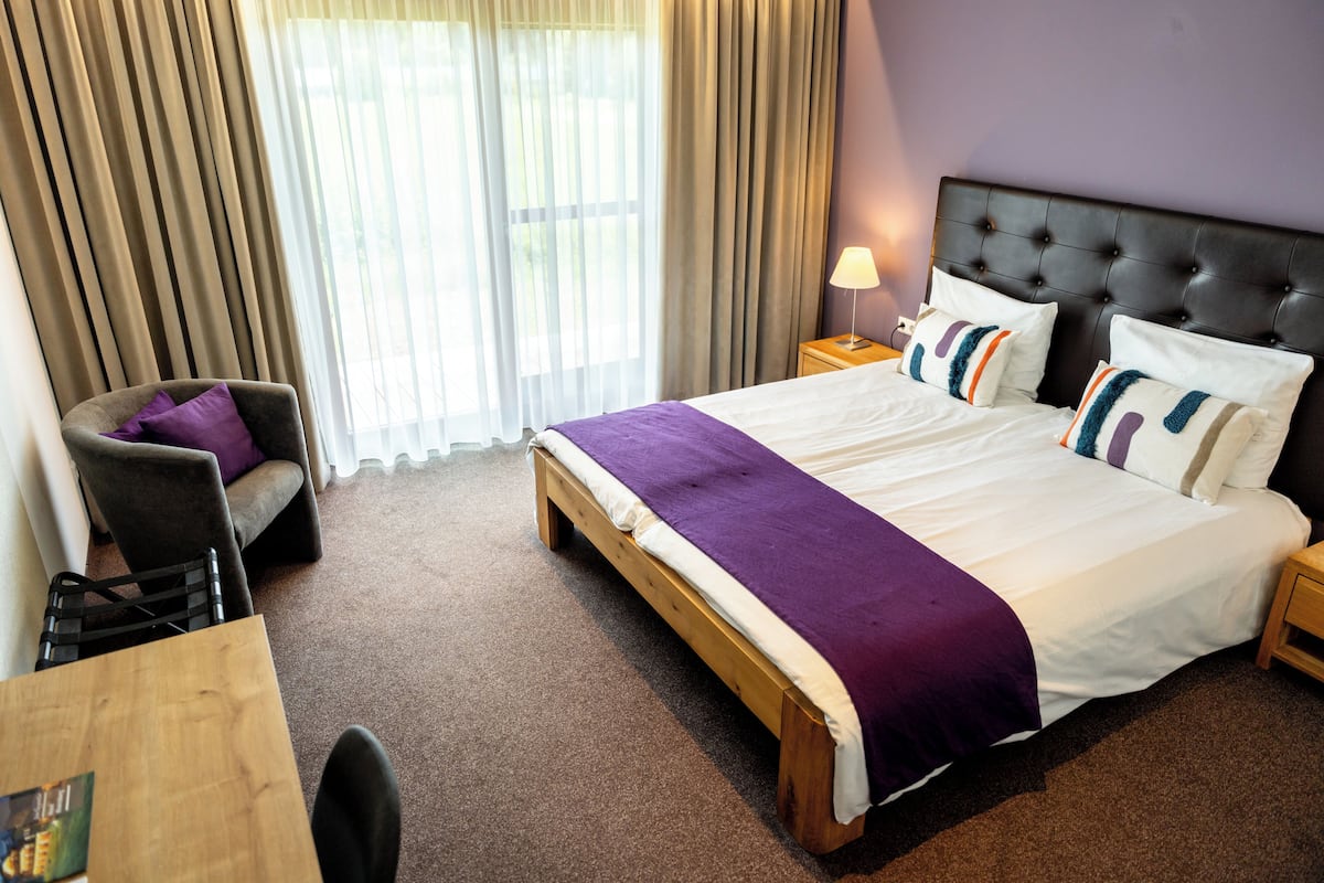 Quarto de hotel com uma cama grande, uma cadeira, uma mesa de madeira e uma mesinha de cabeceira com abajur.