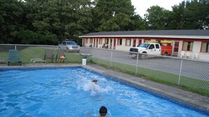 Uma piscina com uma pessoa nadando. Há uma cerca ao redor da piscina e um edifício com telhado vermelho ao fundo.