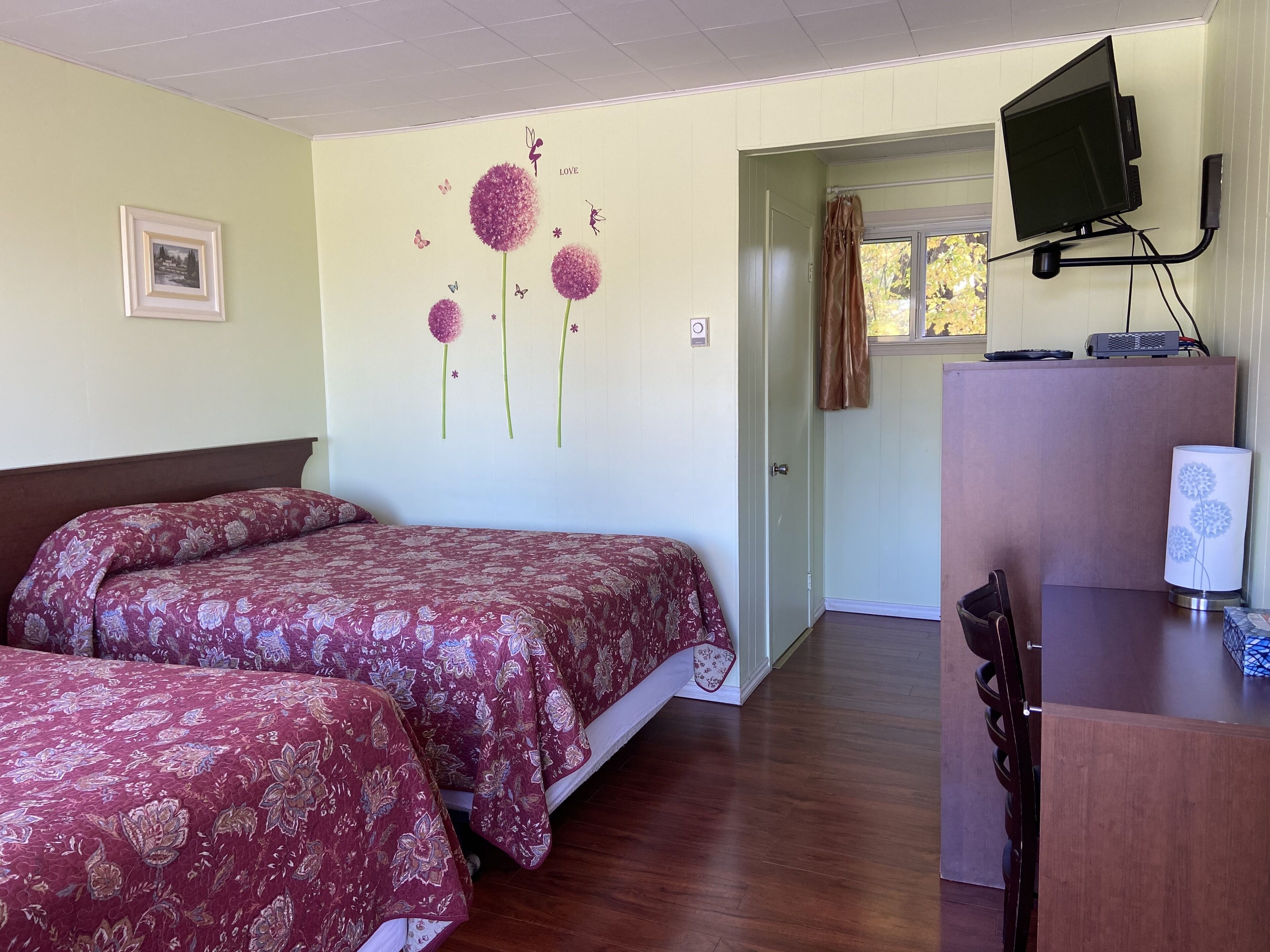 Quarto de hotel com duas camas, uma escrivaninha, televisão e uma parede com decoração floral.