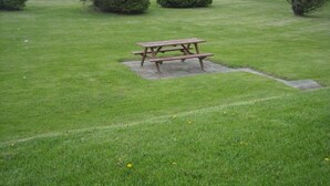 Uma mesa para piquenique em uma área ajardinada, com arbustos bem cuidados e árvores ao fundo.