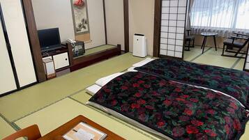Um quarto tradicional japonês com tatames, uma cama com colcha floral, uma escrivaninha com telefone e um caderno, e um futon.