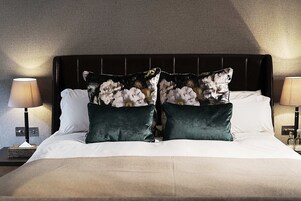 Cama bem arrumada com lençóis brancos e duas almofadas decorativas, com duas luminárias de cabeceira sobre um criado-mudo de madeira escura.