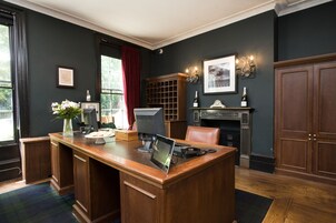 Escritório formal com uma mesa de madeira, computador e telefone. Há uma estante de livros, uma lareira e uma janela grande com cortinas.