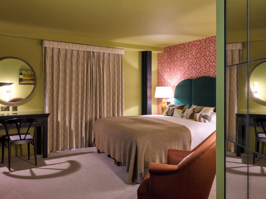 Quarto de hotel com cama, escrivaninha, cadeira, espelho e papel de parede estampado.