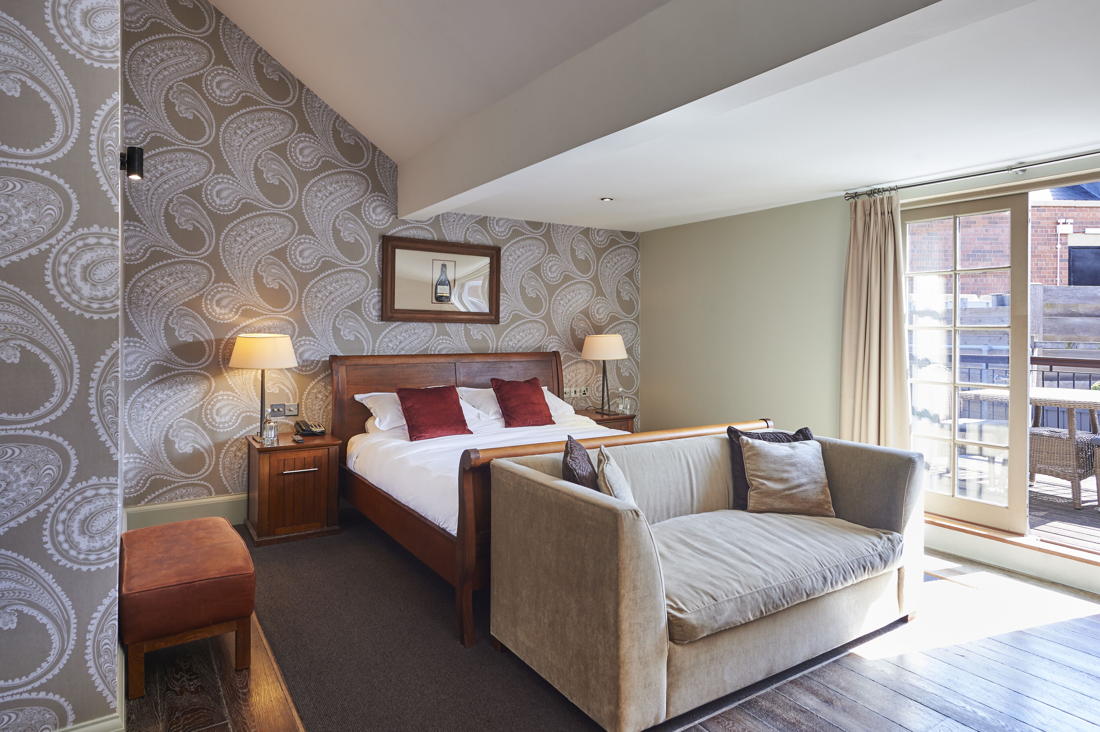 Quarto de hotel com cama, duas mesinhas de cabeceira com luminárias, um sofá, uma janela com cortinas e papel de parede estampado.