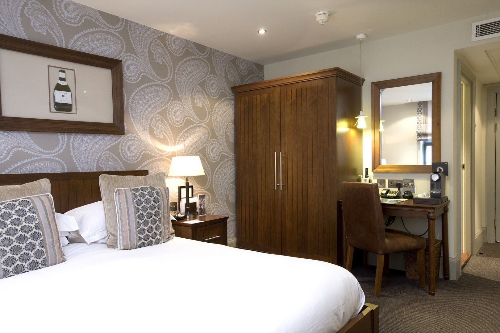Quarto de hotel com cama, escrivaninha, cadeira, abajur, espelho e guarda-roupa.