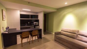 Um espaço compacto com cozinha compacta, duas cadeiras de vime, um sofá e uma parede verde de destaque.