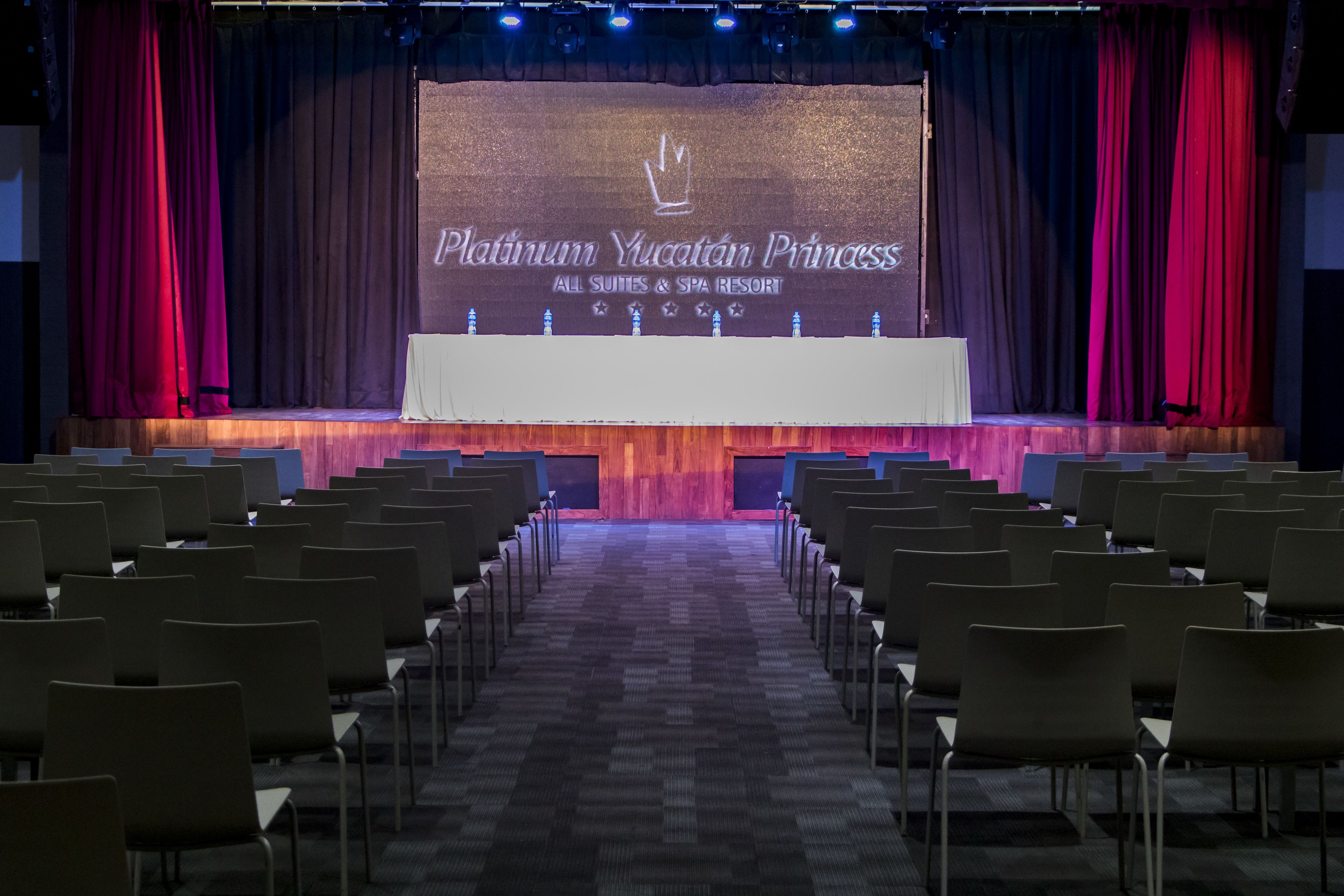 Uma sala de conferências com fileiras de cadeiras voltadas para um palco com uma tela grande exibindo "Platinum Yucatán Princess All Suites & Spa Resort".