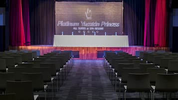 Uma sala de conferências com fileiras de cadeiras voltadas para um palco com uma tela grande exibindo "Platinum Yucatán Princess All Suites & Spa Resort".