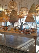 Um buffet de café da manhã com luminárias de vime pendentes, diversas opções de pães e doces, e piso de azulejos.