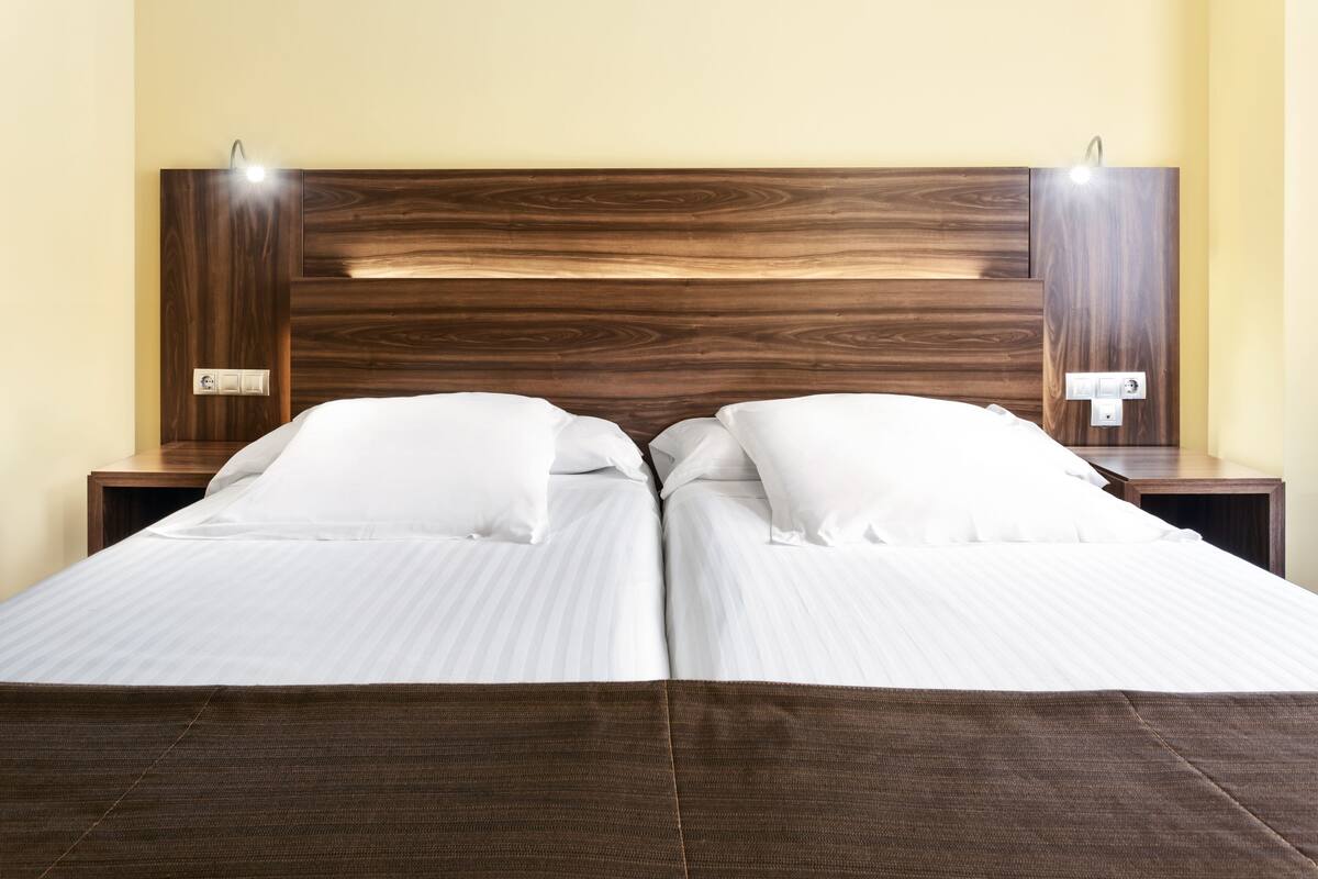 Uma cama de casal com cabeceira de madeira, duas mesas de cabeceira e um candeeiro fixo na parede.
