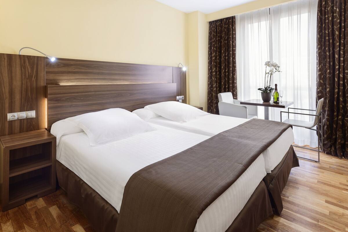 Quarto de hotel com cama grande, mesas de cabeceira, escrivaninha com cadeira e vista do exterior através de cortinas translúcidas.