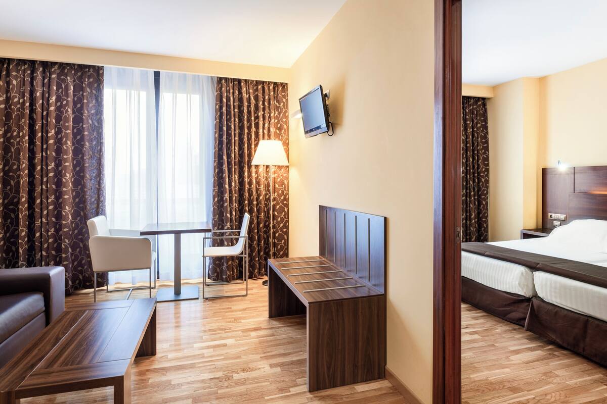 Quarto de hotel com uma cama, uma escrivaninha com cadeira, uma TV fixada na parede e uma janela com cortinas.
