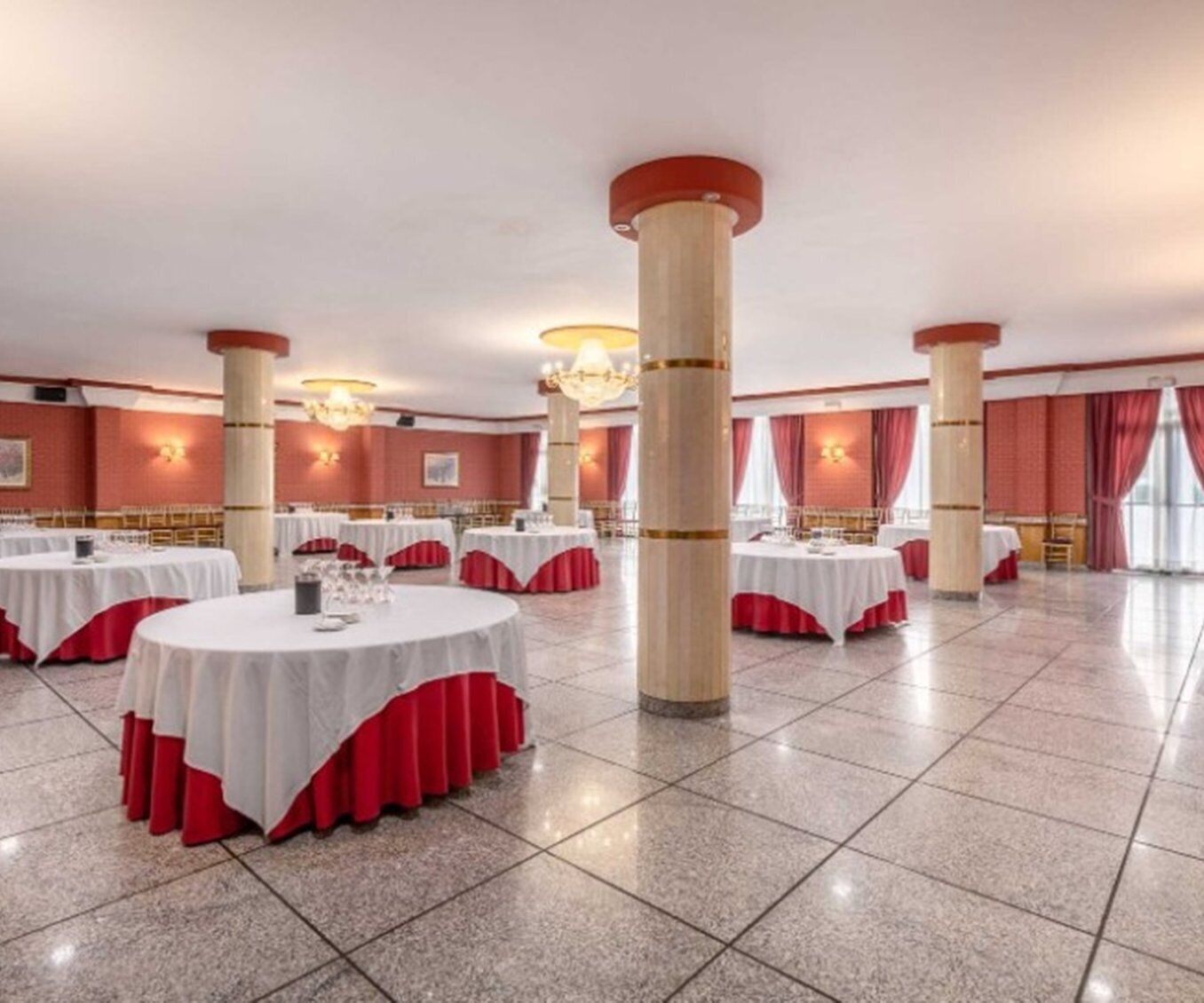 Uma sala de banquetes com mesas redondas, toalhas vermelhas e brancas e colunas imponentes.