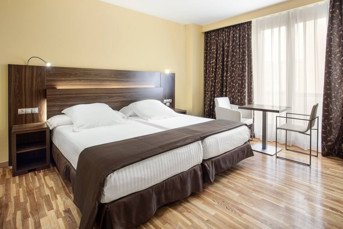 Quarto de hotel com uma cama grande, mesa de cabeceira, escrivaninha com cadeira e janela com cortinas.