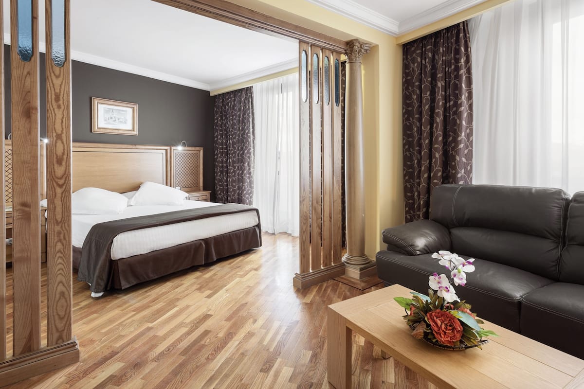 Quarto de hotel com uma cama, um sofá, piso de madeira, janela com cortinas e um quadro pendurado na parede.