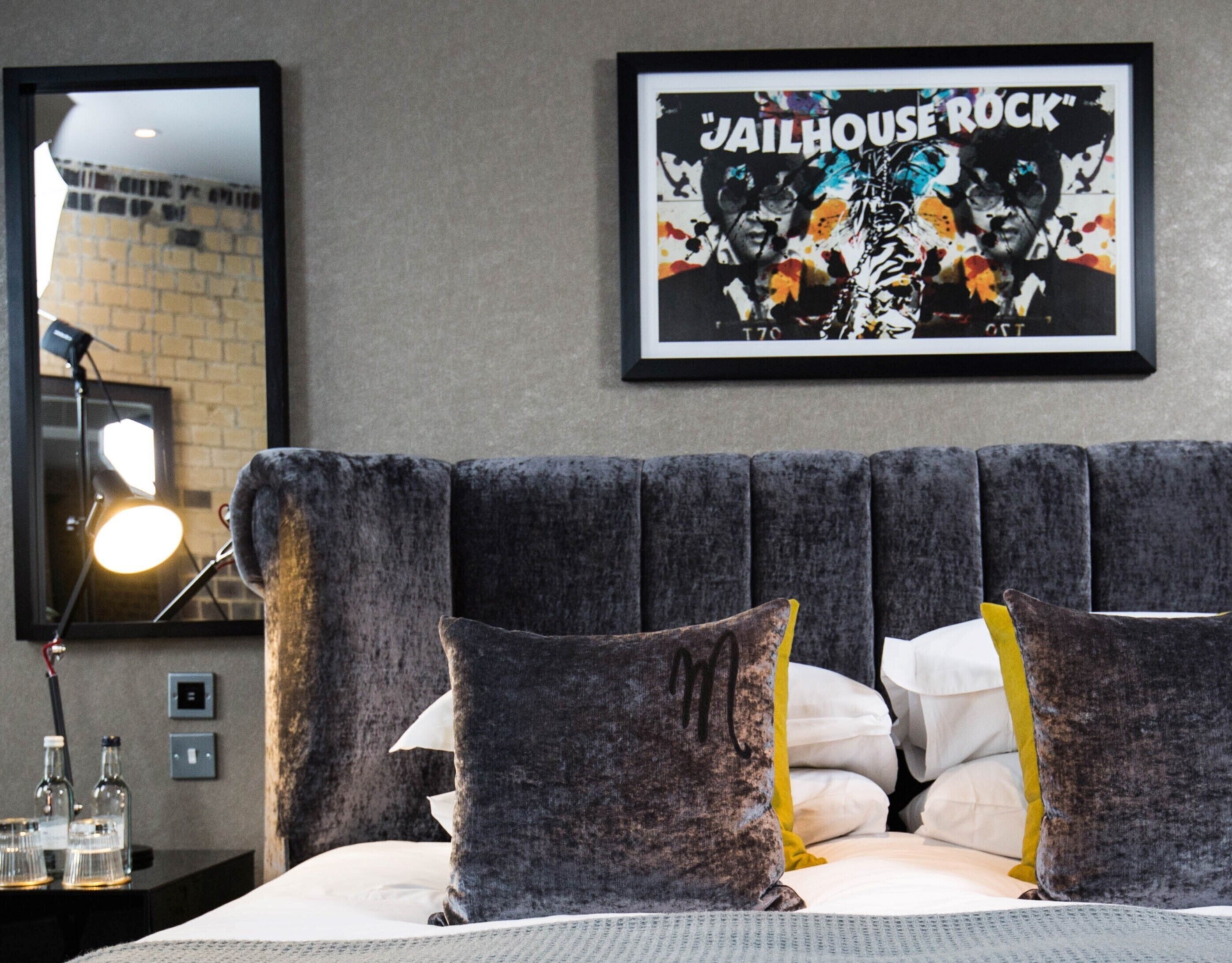 Uma cama com cabeceira escura, um quadro na parede e uma luminária em uma mesa de cabeceira.