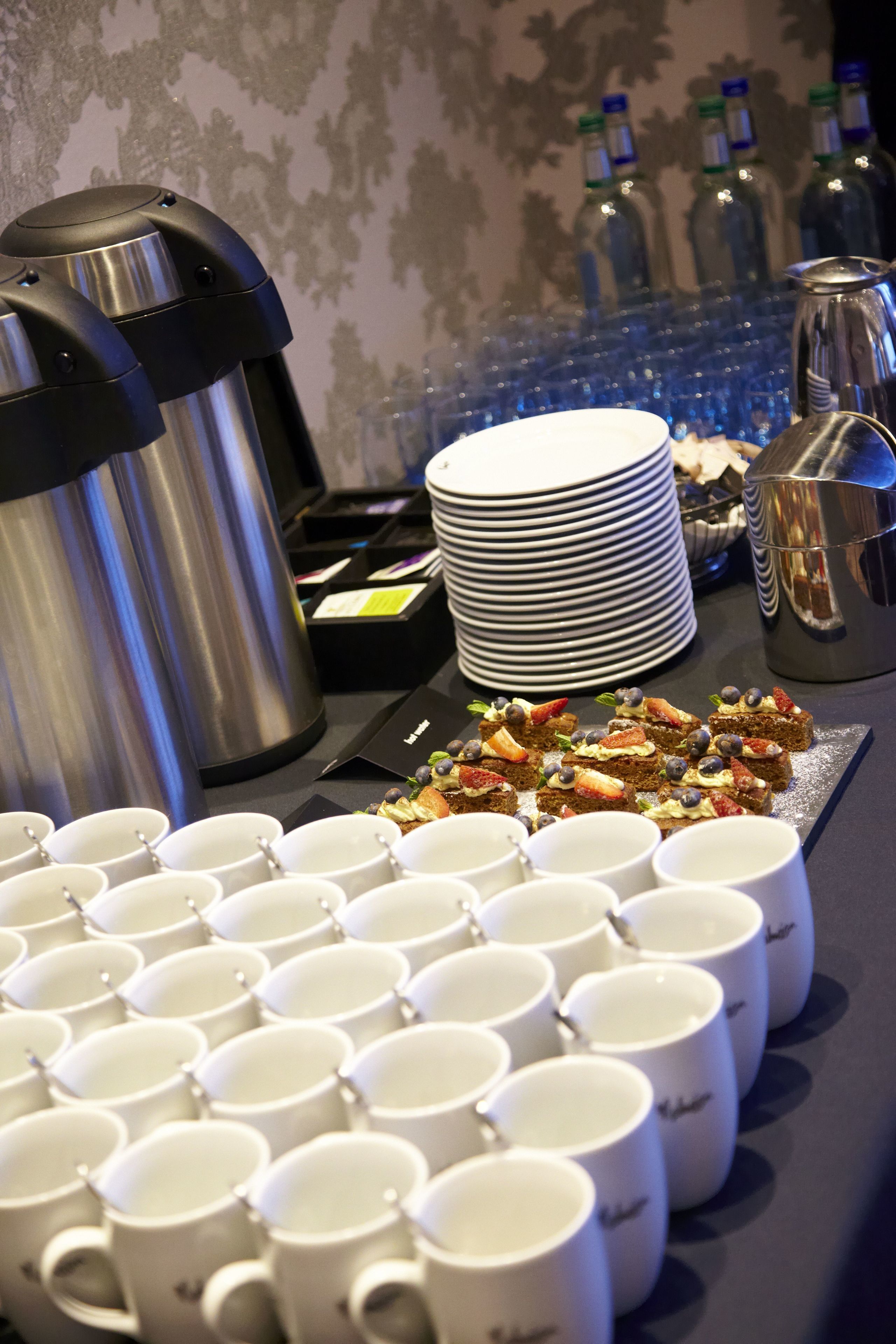 Disposição de buffet com comida, cafeteiras e xícaras brancas empilhadas.