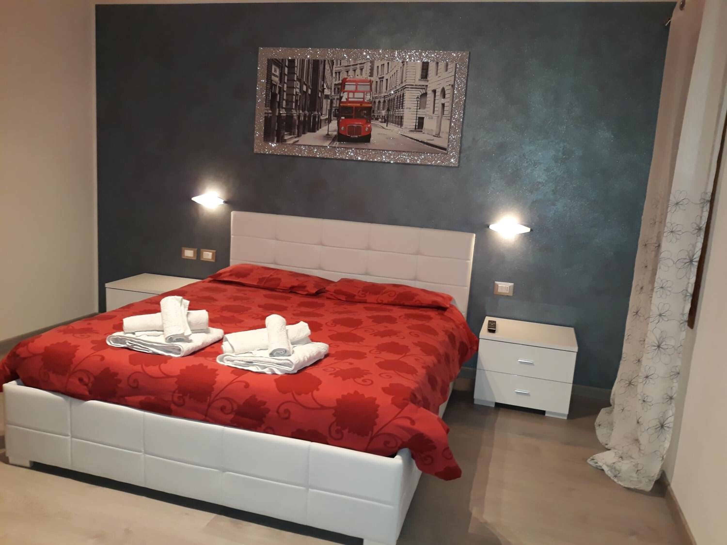 Cama bem arrumada com roupa de cama vermelha e cabeceira branca, duas mesas de cabeceira com abajures, um quadro na parede e cortina com padrão floral.