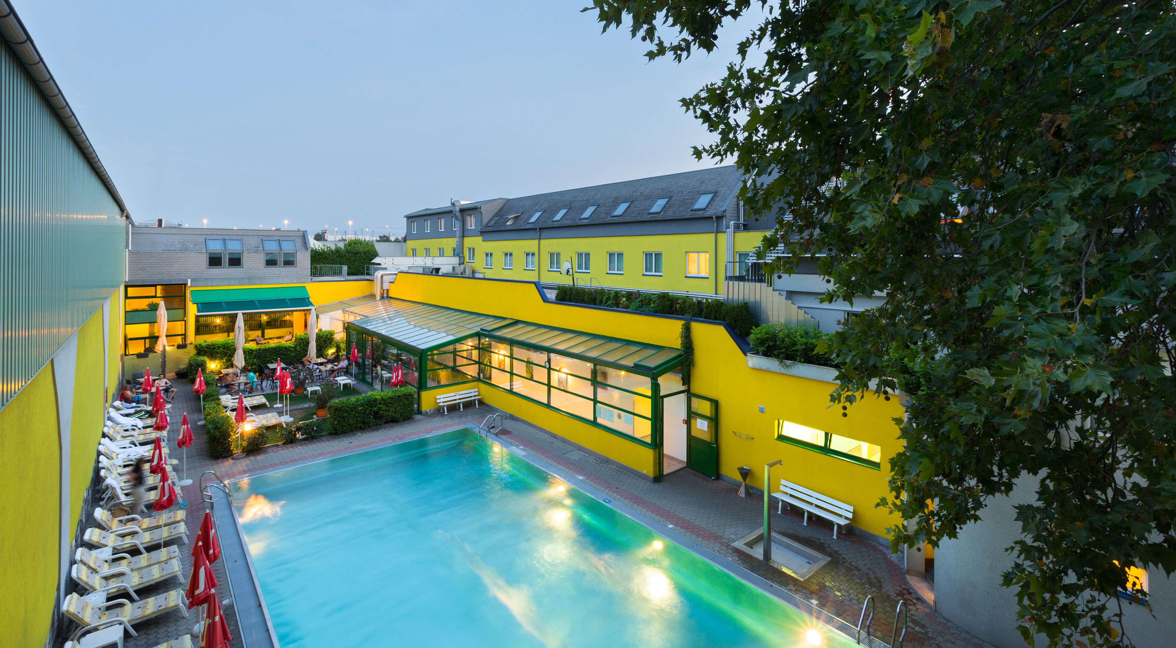 Piscina com espreguiçadeiras e guarda-sóis, área de estar coberta e um edifício com fachada amarela.