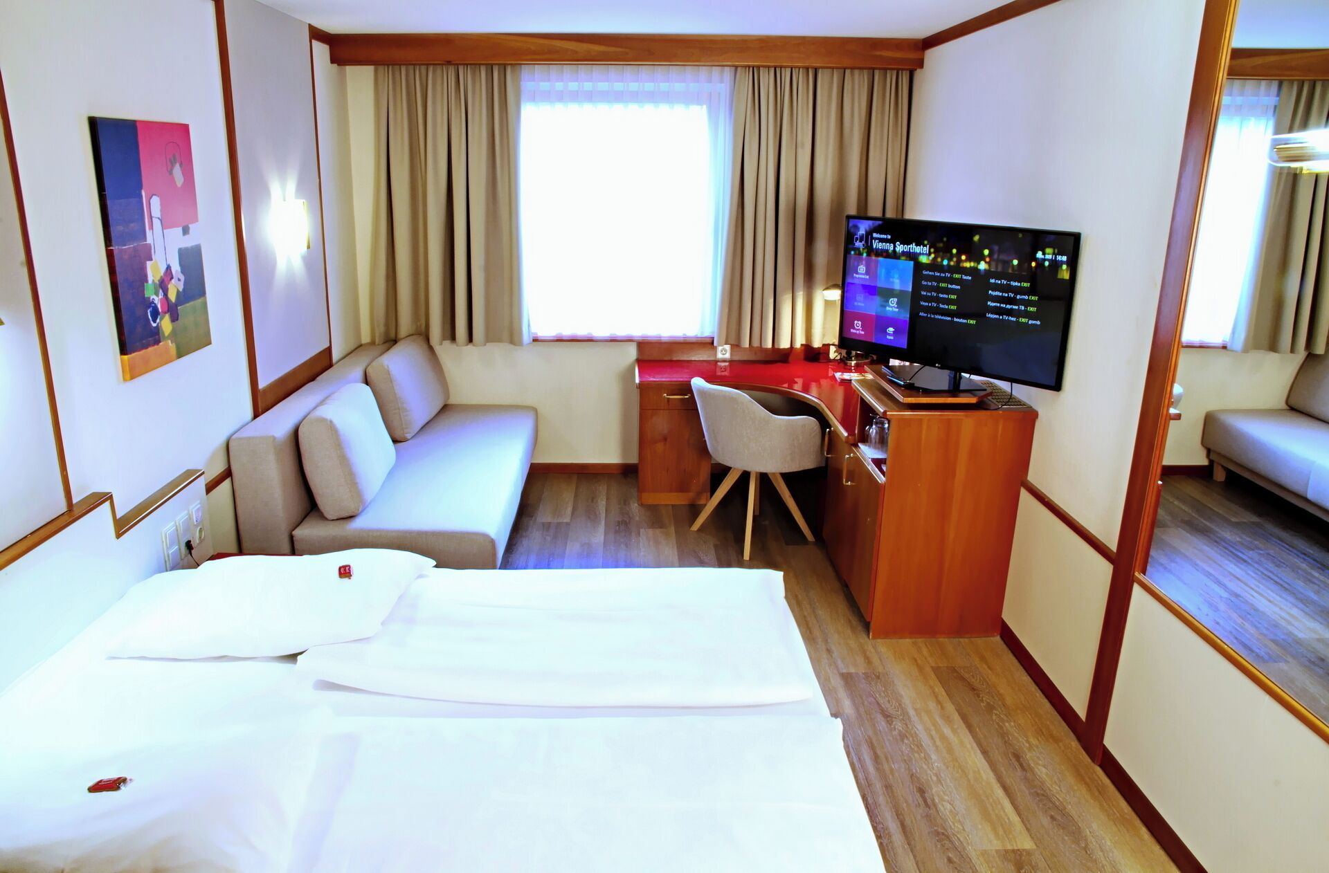 Quarto de hotel com cama, sofá, escrivaninha com computador, televisão e janela com cortinas.