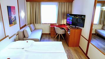 Quarto de hotel com cama, sofá, escrivaninha com computador, televisão e janela com cortinas.