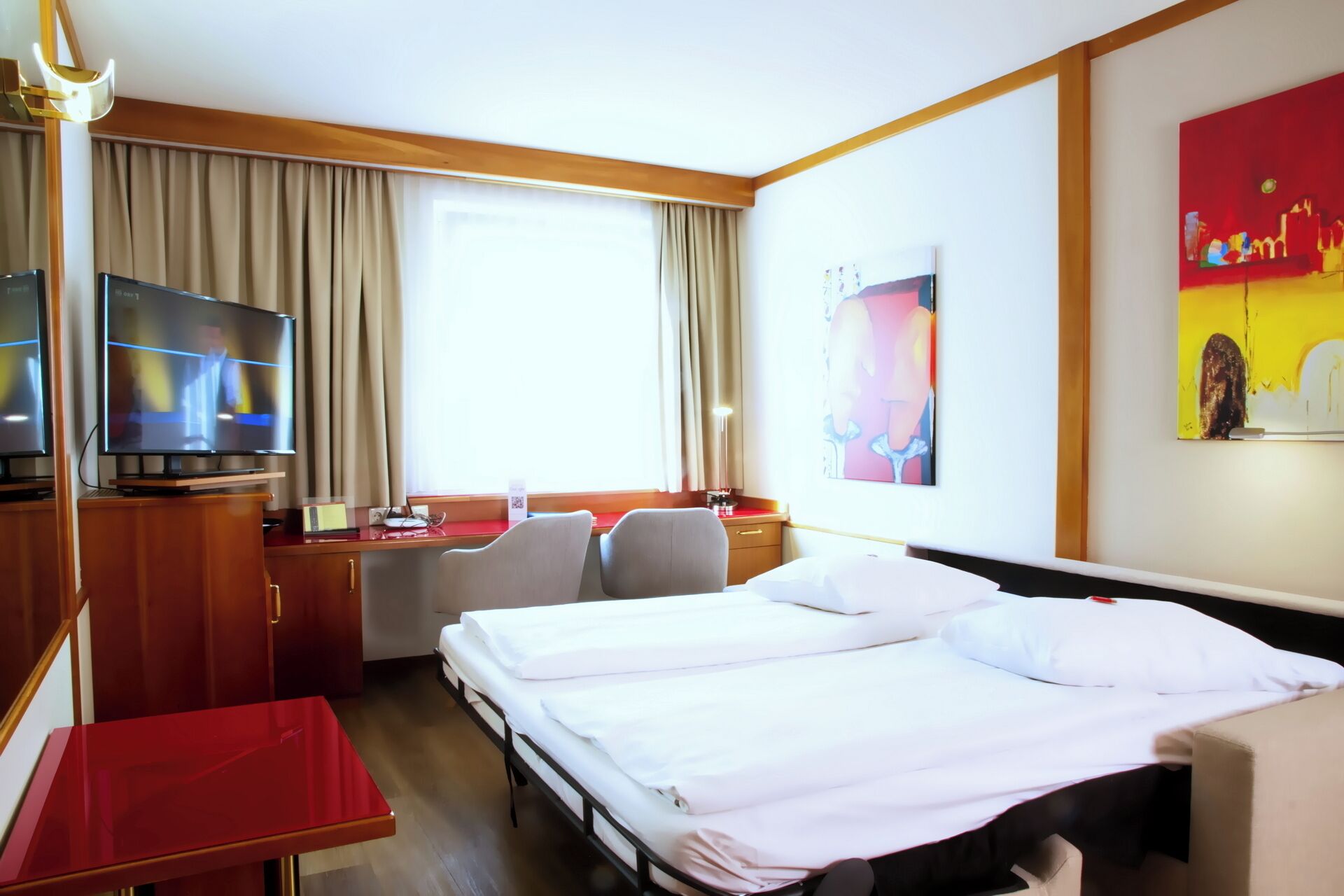 Quarto de hotel com duas camas, uma mesa com cadeira, televisão e janela com cortinas.