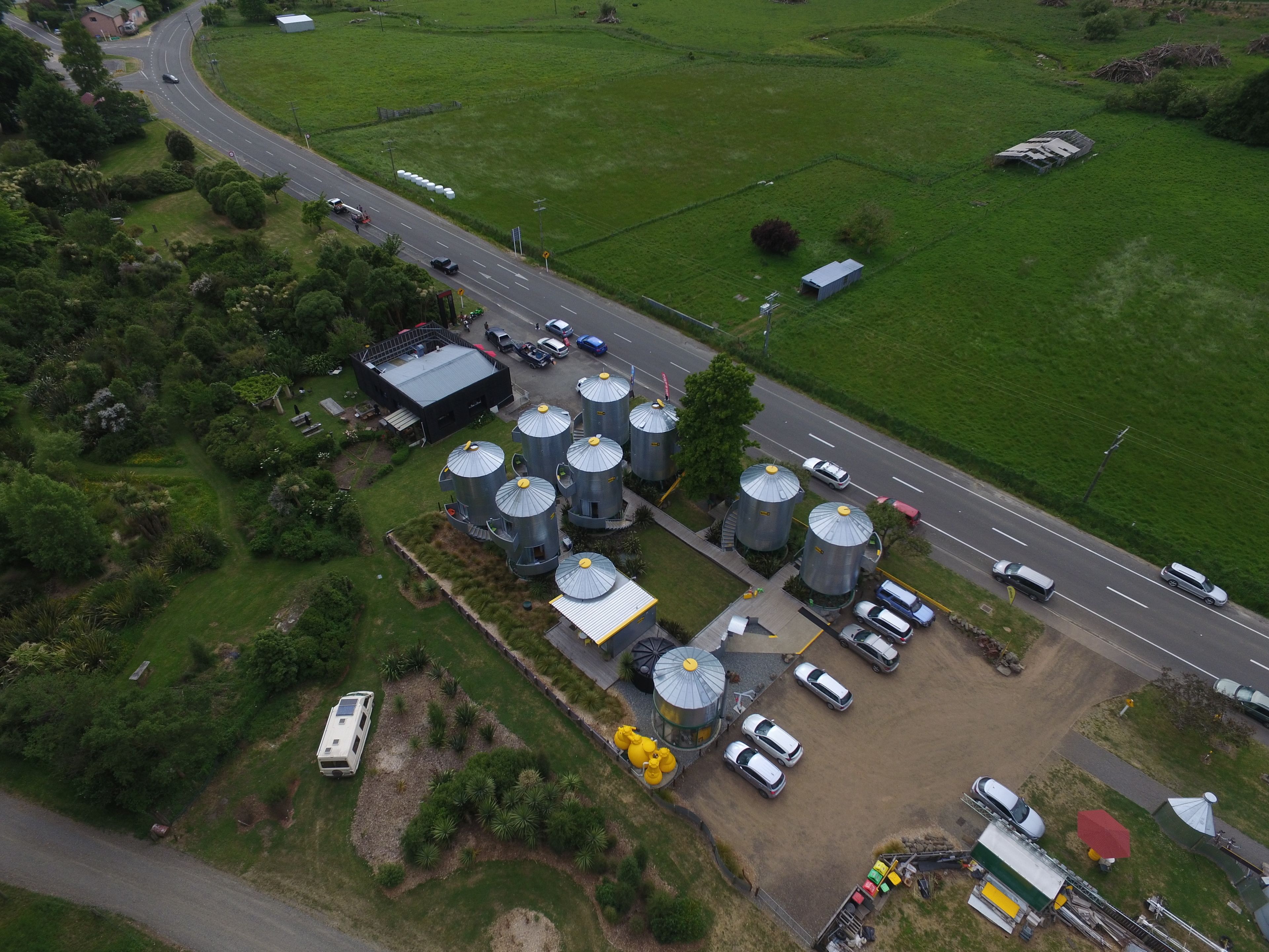 Vista aérea de uma área rural com estrada, campos verdes e vários silos cilíndricos grandes.