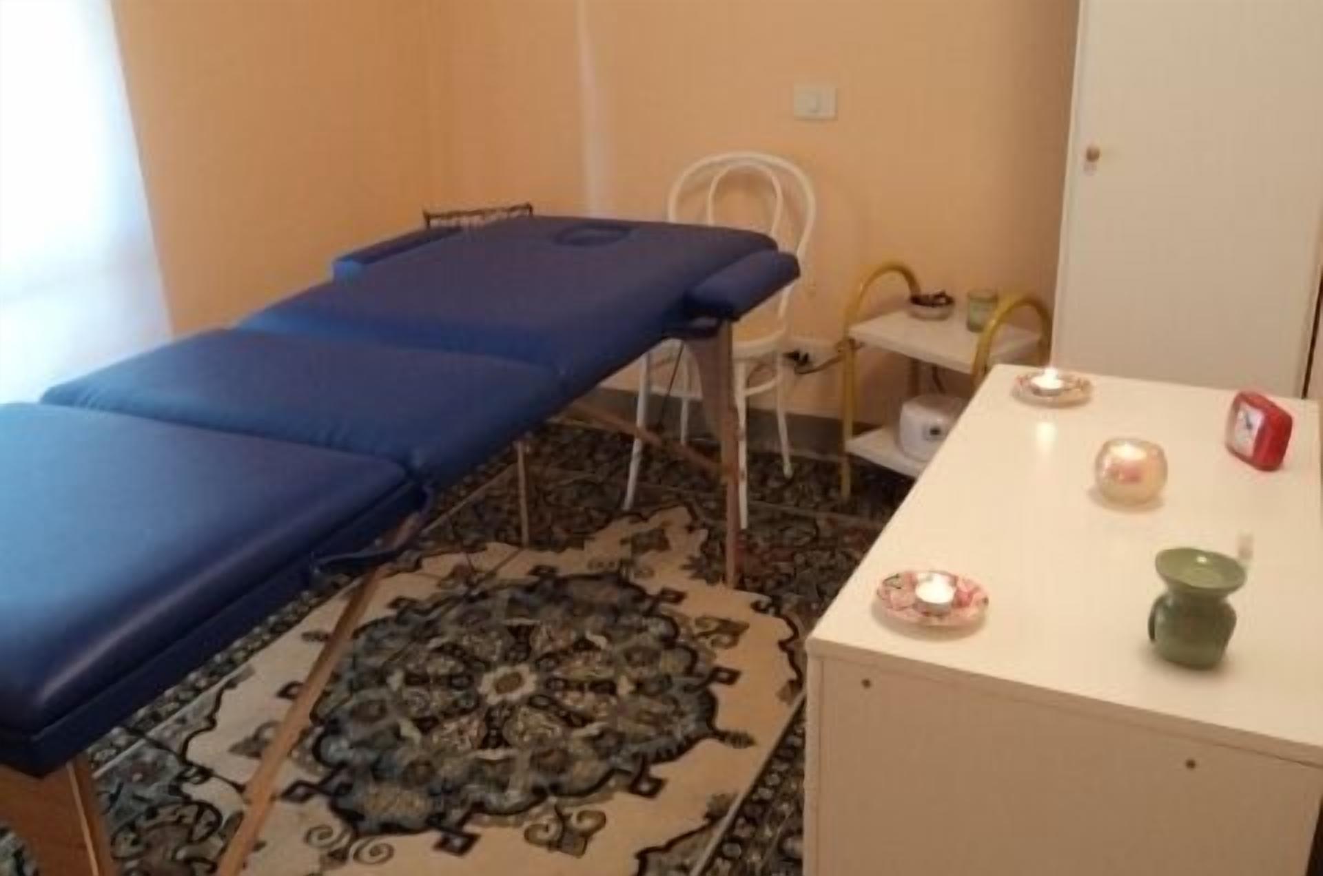 Um quarto com uma mesa de massagem azul, um tapete estampado, uma mesinha branca com velas e uma cadeira branca.