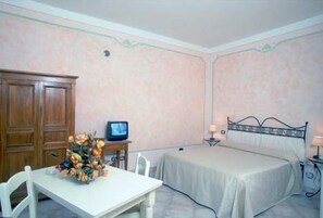 Quarto com cama, televisão sobre uma mesinha, escrivaninha com um vaso de flores e guarda-roupa.