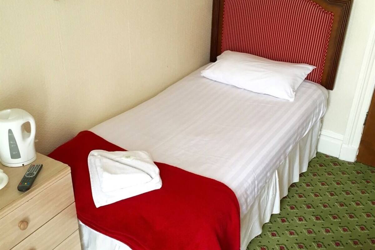Cama bem arrumada com cabeceira vermelha, travesseiro branco e manta branca. Um criado-mudo de madeira com uma chaleira branca e um controle remoto. O quarto possui um tapete verde estampado e um quadro na parede.
