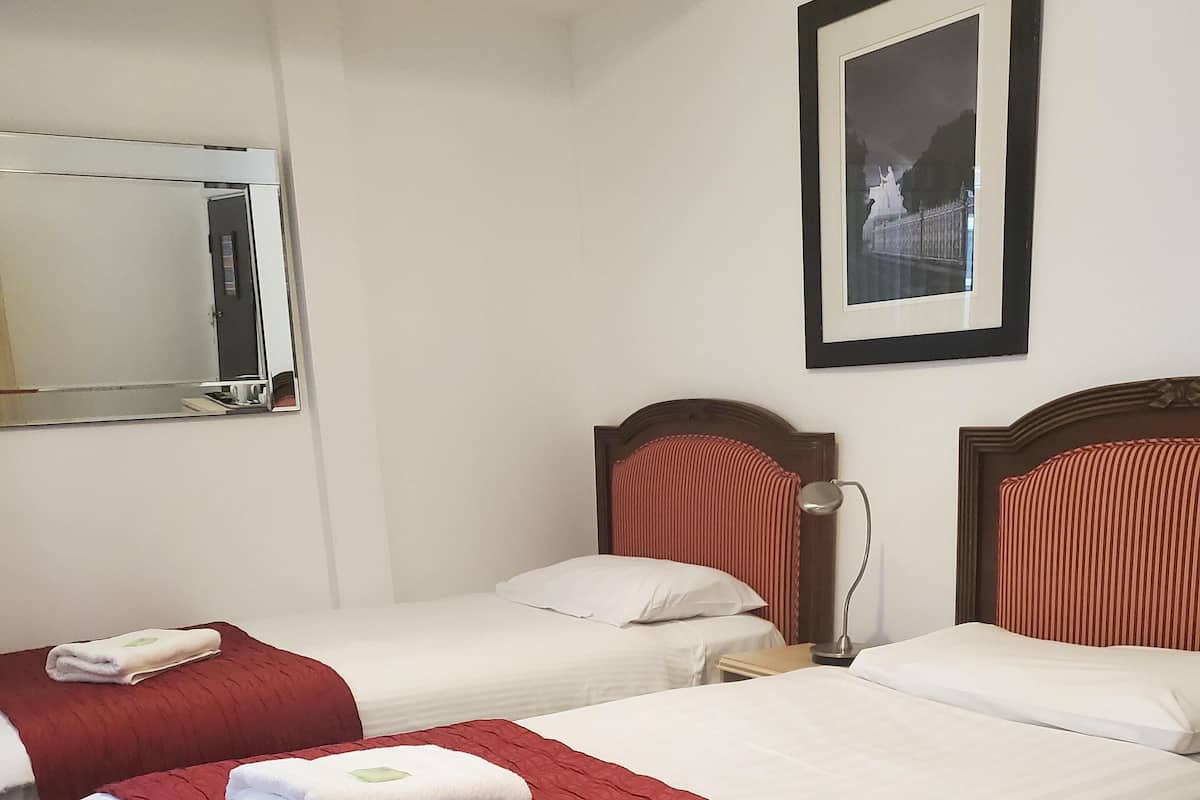 Quarto de hotel com duas camas de solteiro, cada uma com cabeceira vermelha e roupa de cama branca. Há uma moldura com uma imagem na parede, acima das camas.