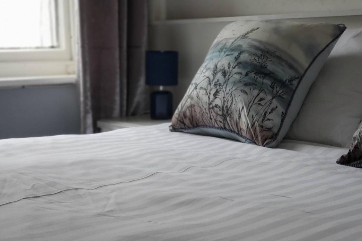 Cama bem arrumada com travesseiro floral, luminária de cabeceira e janela com cortinas.