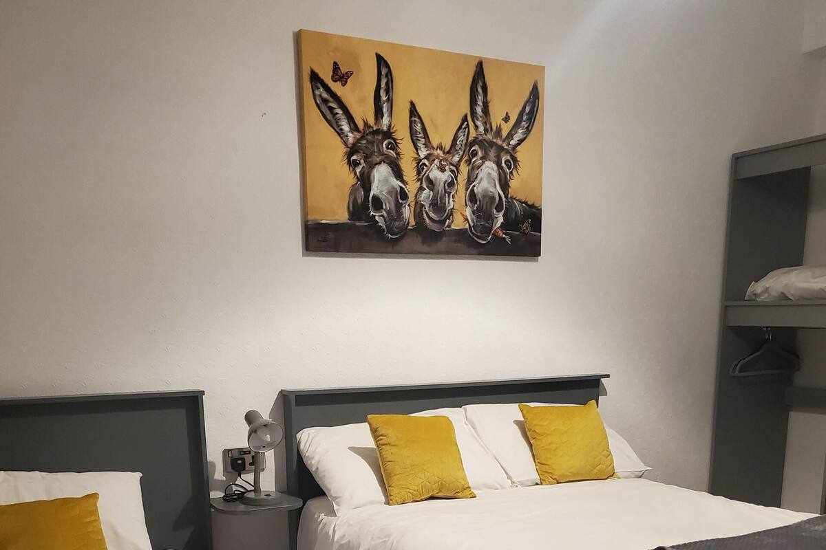 Quarto de hotel com duas camas, cada uma com almofadas brancas e mostarda, um quadro de burros na parede e uma pequena mesa com uma luminária.
