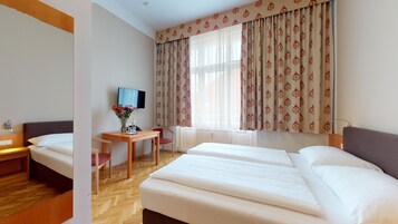 Quarto de hotel com duas camas, uma mesa com cadeira, televisão e janela com cortinas estampadas.