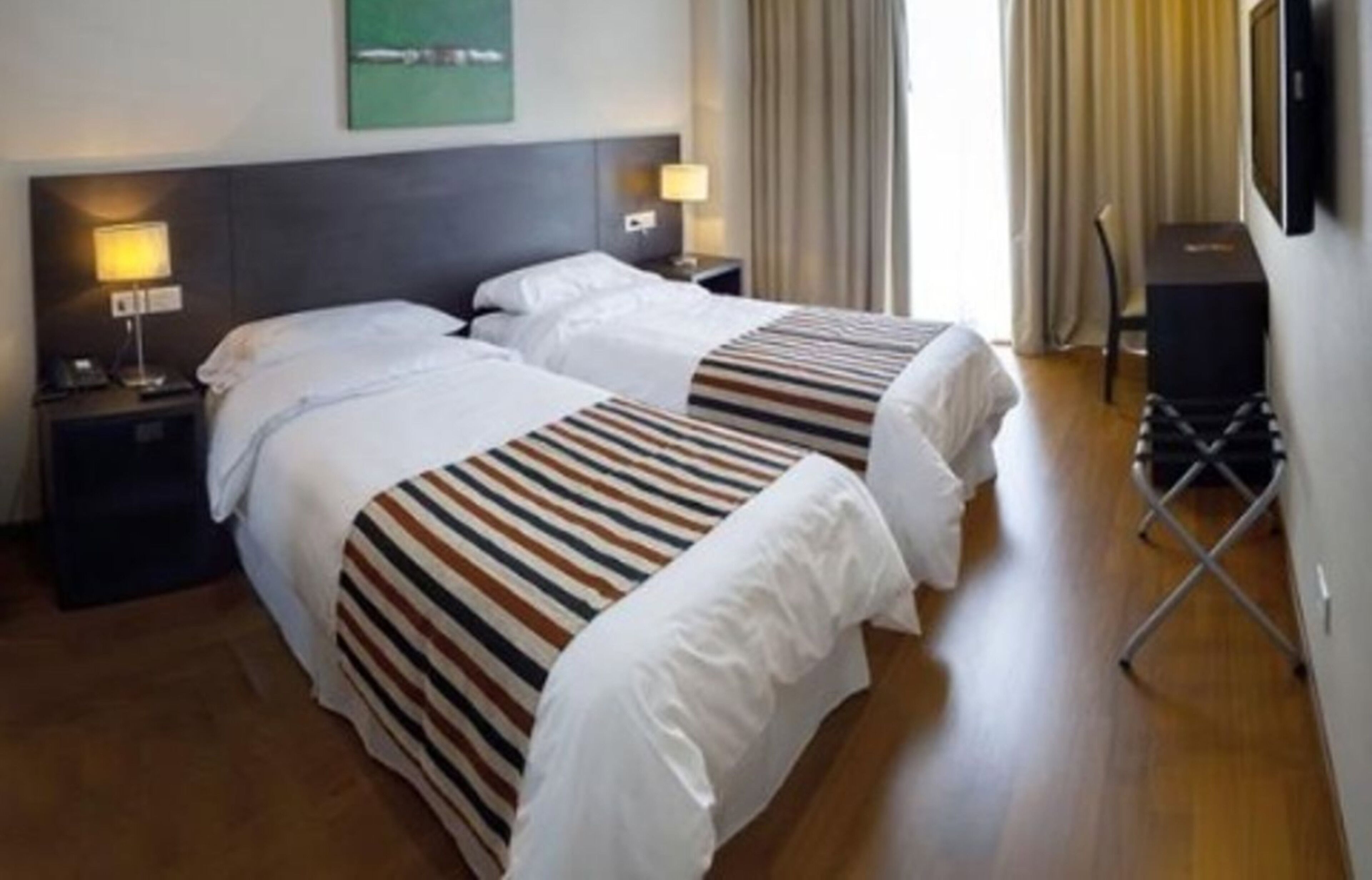 Quarto de hotel com duas camas, uma escrivaninha, uma cadeira e uma televisão na parede.