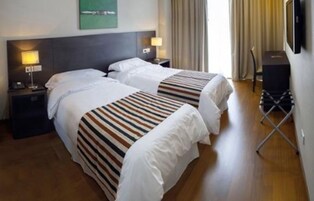 Quarto de hotel com duas camas, uma escrivaninha, uma cadeira e uma televisão na parede.
