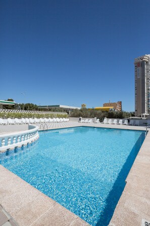 Uma piscina retangular com água azul cristalina, rodeada por cadeiras de descanso brancas e um deck revestido de azulejos.