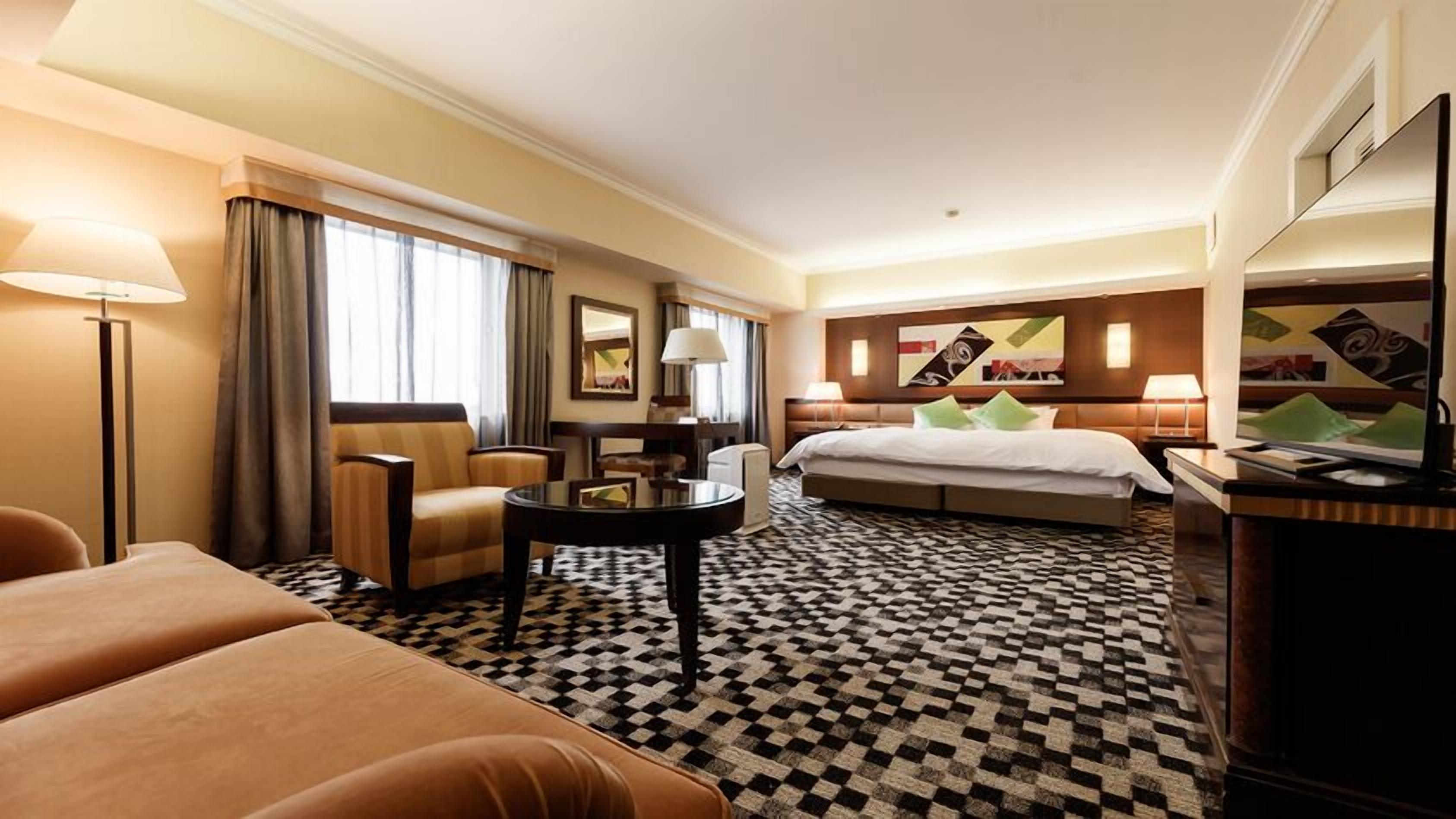 Quarto de hotel com cama, duas poltronas, escrivaninha, abajur e chão xadrez.