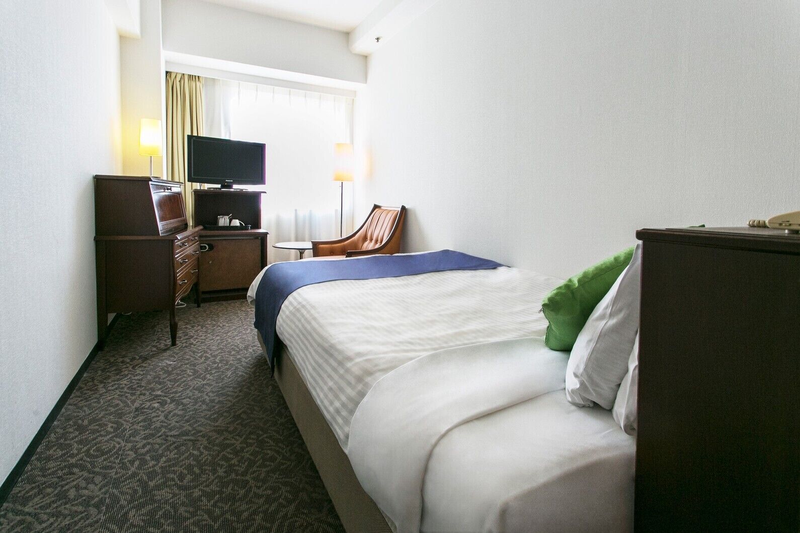 Quarto de hotel com cama, escrivaninha com televisão, uma cadeira e cómoda.