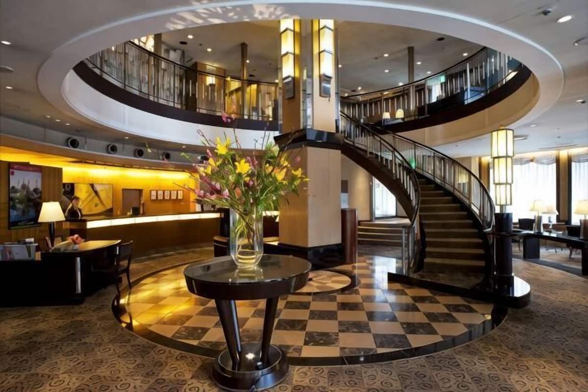 Um saguão de hotel com uma escadaria central, um balcão de recepção e um vaso decorativo.