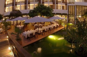Uma área de refeições ao ar livre com mesas e guarda-sóis, piscina e um edifício com vários andares e grandes janelas.
